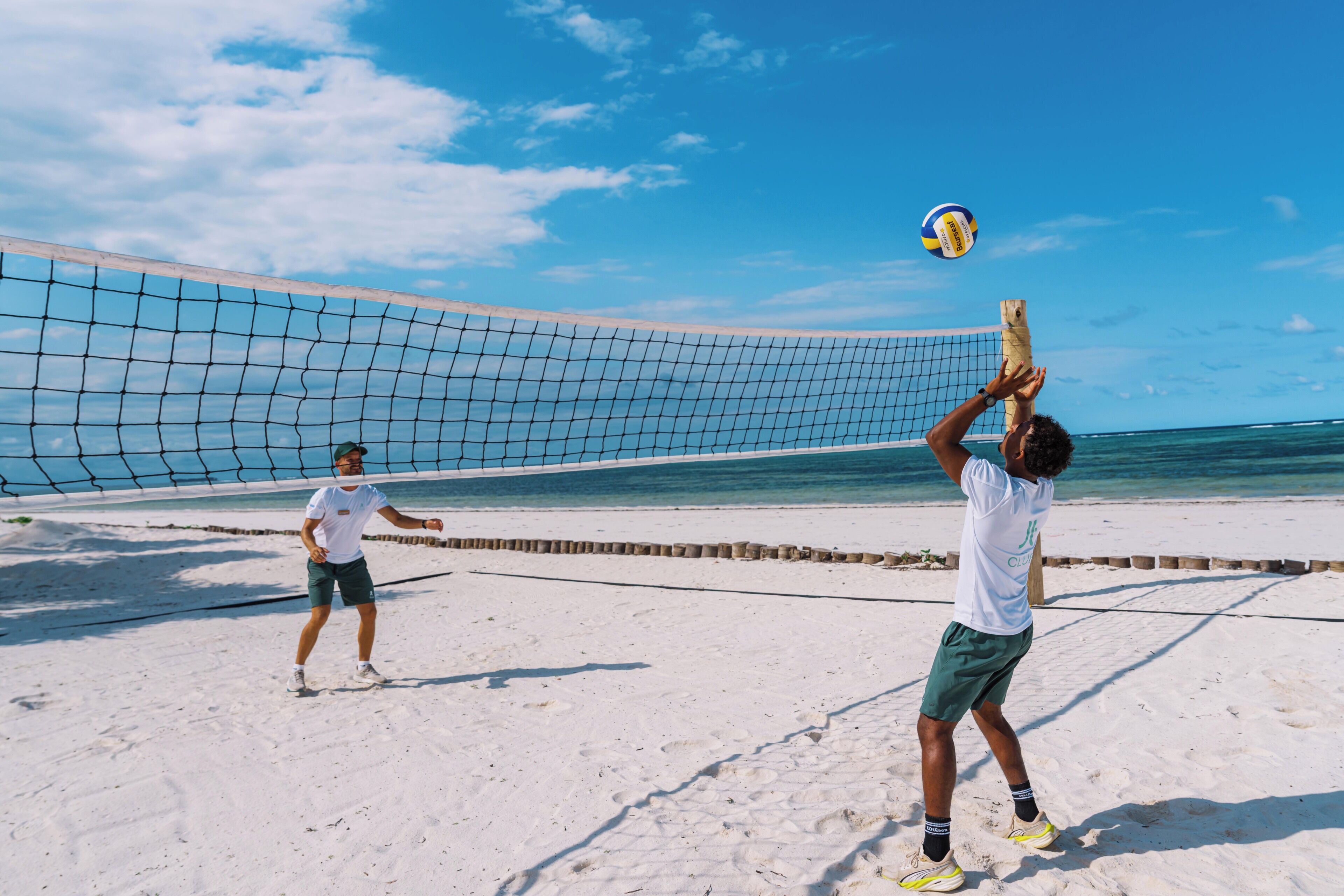 Jogo de voleibol de praia com rede e jogadores em quadra de areia.