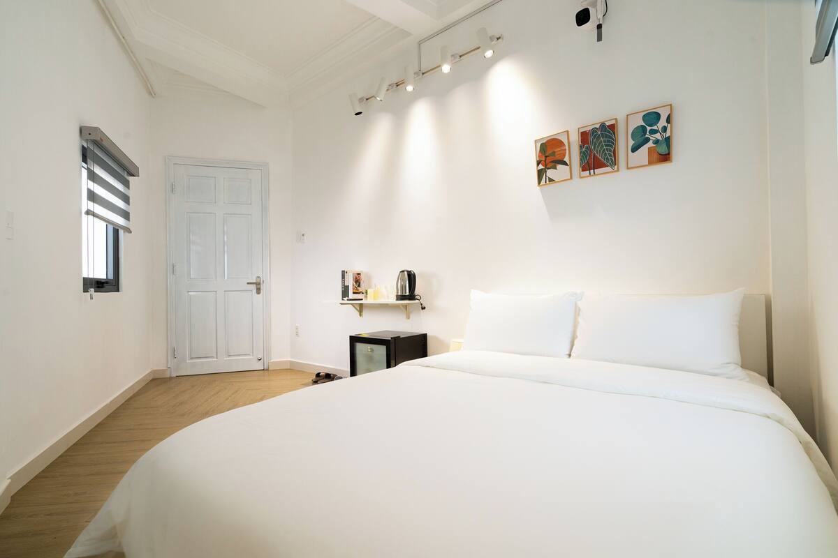 Quarto com uma cama grande, uma porta branca, uma mesinha de cabeceira com abajur e uma janela com persianas.