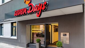 Um hotel chamado 'Seven Days' com fachada moderna, amplas janelas e duas plantas em vasos na frente.