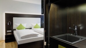 Quarto de hotel moderno com piso de madeira, uma cama grande com roupa de cama branca e almofadas verdes, uma mesa de cabeceira e uma pia embutida com uma toalha amarela.