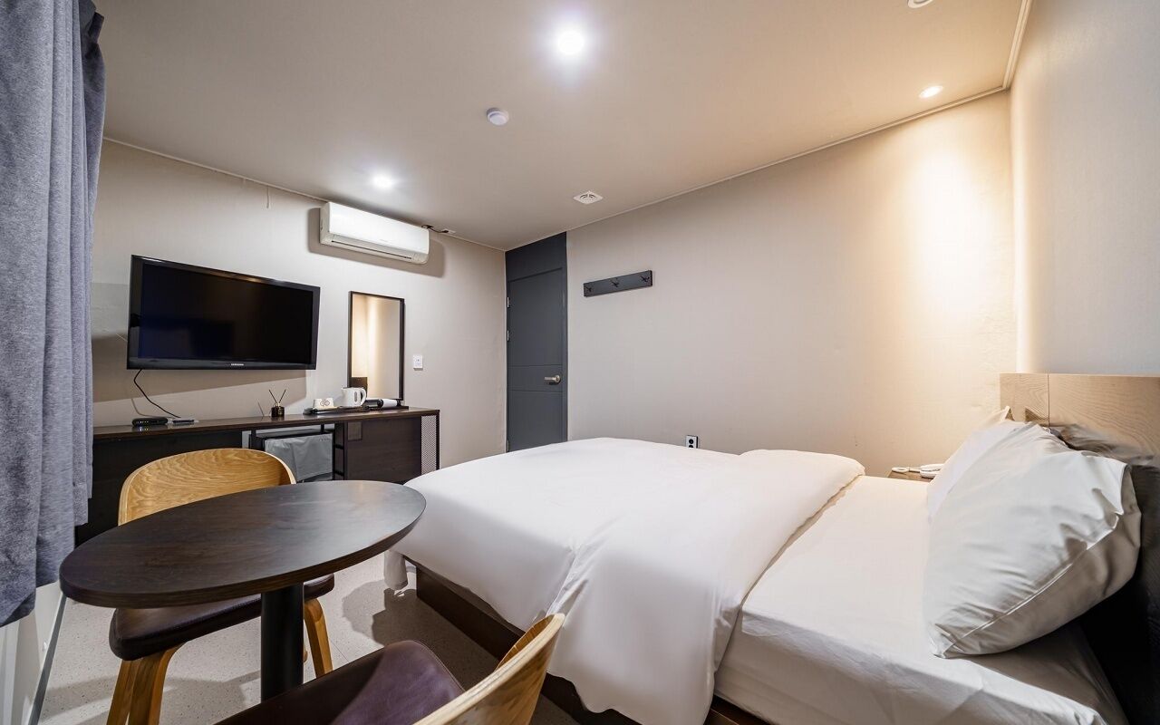 Quarto de hotel com uma cama, uma escrivaninha com computador, uma televisão e uma cadeira.