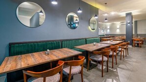 Um restaurante com mesas de madeira e bancos estofados verdes, luminárias pendentes e espelhos redondos nas paredes.