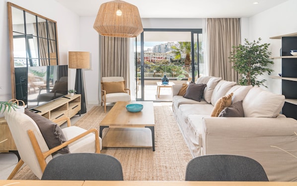 Uma sala de estar moderna com um sofá, cadeiras, mesa de centro, TV e varanda com plantas.