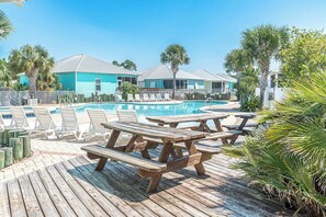 Área de piscina com mesas de piquenique de madeira e cadeiras de lounge, cercada por palmeiras e um edifício com fachada azul.