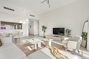 Uma sala de estar moderna com televisão de tela plana, área de refeições e cozinha ao fundo, além de uma grande janela que permite entrada de luz natural.