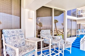 Varanda com cadeiras brancas e uma mesa, janelas amplas com vista para palmeiras e um tapete azul.