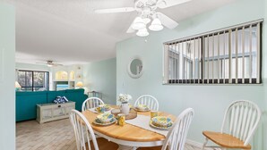 Uma área de refeições com mesa de madeira posta para quatro pessoas, cadeiras brancas e ventilador de teto.
