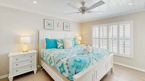 Quarto com cama branca, roupa de cama verde-água, ventilador de teto e janela com persianas.