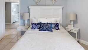 Quarto com cama grande, duas mesas de cabeceira, cabeceira branca e peças decorativas na parede.
