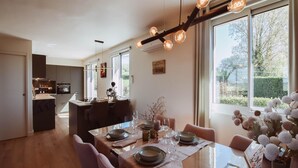 Uma sala de jantar moderna com uma mesa de madeira posta para quatro pessoas, cozinha ao fundo e grandes janelas com vista para o verde.
