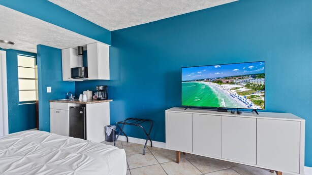 Quarto com uma cama, televisão mostrando uma vista da praia, cozinha compacta e uma janela.
