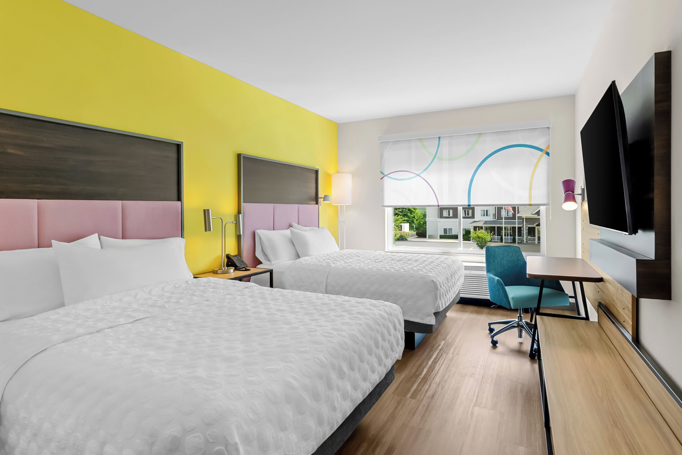 Quarto de hotel com duas camas, uma escrivaninha, uma cadeira, televisão e janela com vista para edifícios.