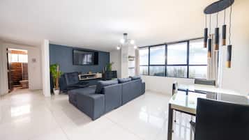 Uma sala de estar moderna com um sofá cinzento, uma TV fixada na parede e um espaço de jantar com mesa e cadeiras.