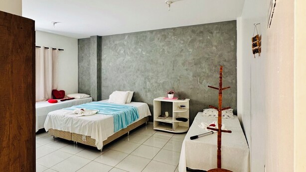 Quarto de hotel moderno com duas camas, uma cadeira de madeira pequena, uma mesinha de cabeceira e uma parede cinza texturizada.