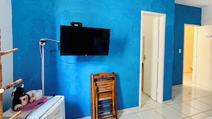 Quarto com parede azul, televisão de tela plana montada acima de uma geladeira branca, um banquinho de madeira e uma porta branca que dá para outro cômodo.