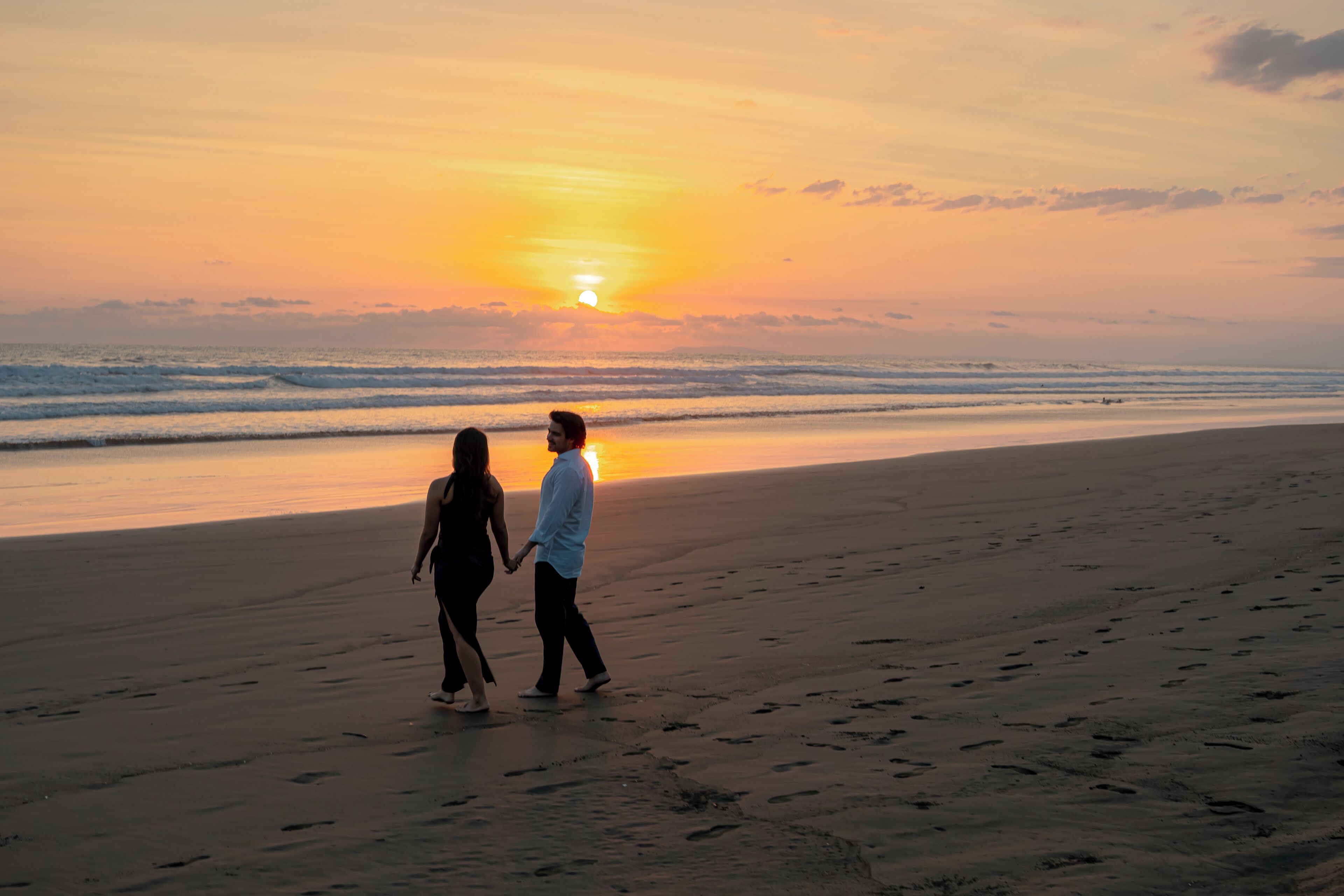 Uma praia com pegadas, um pôr do sol e um casal caminhando.