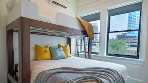 Uma cama beliche com estrutura de madeira, roupa de cama branca e vista da cidade através de grandes janelas.