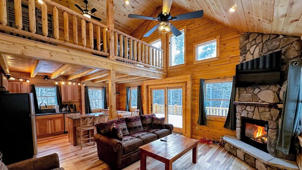 Uma cabana espaçosa com interior em madeira, lareira de pedra, área de refeições e uma sala de estar aconchegante com sofá e mesa de centro.