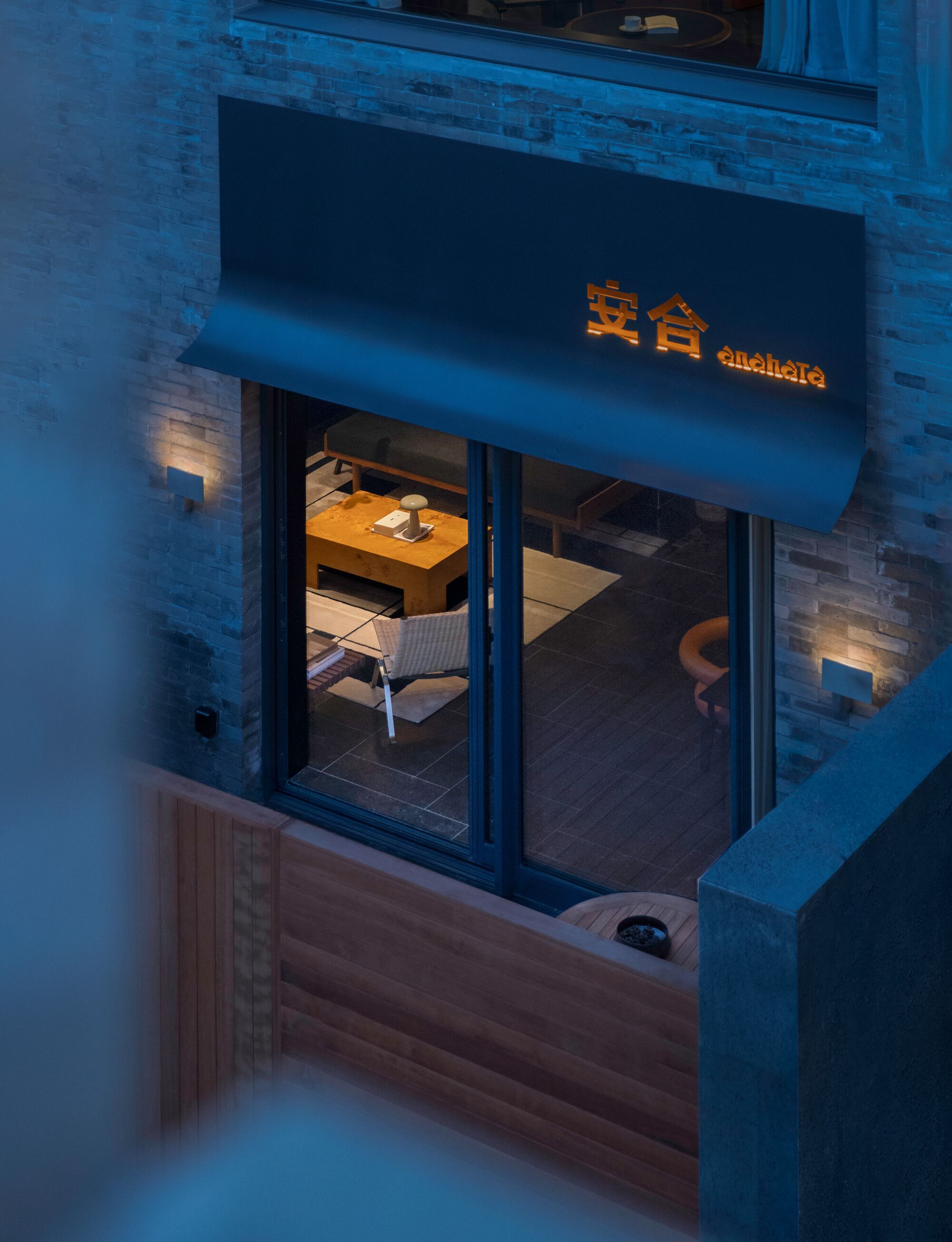 Um edifício moderno com área visível de restaurante ou café, apresentando uma mesa e cadeiras de madeira, um banco e uma pequena mesa com abajur.