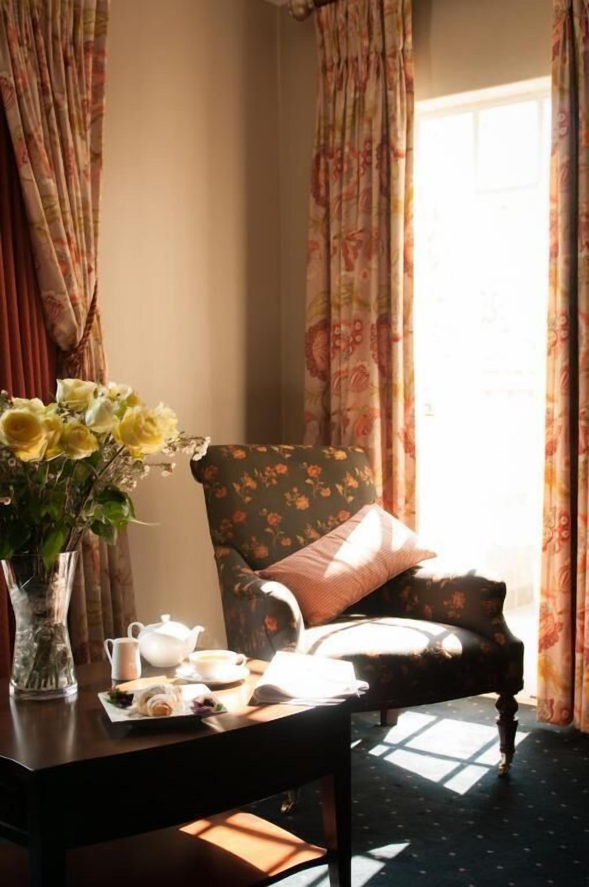 Um quarto com uma poltrona de padrão floral, um vaso com flores amarelas, uma mesa com bule, xícara e pires, e uma janela com luz do sol.