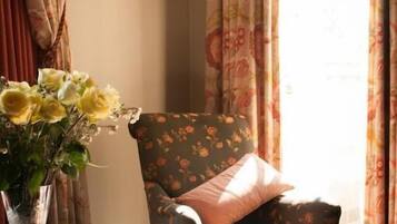 Um quarto com uma poltrona de padrão floral, um vaso com flores amarelas, uma mesa com bule, xícara e pires, e uma janela com luz do sol.