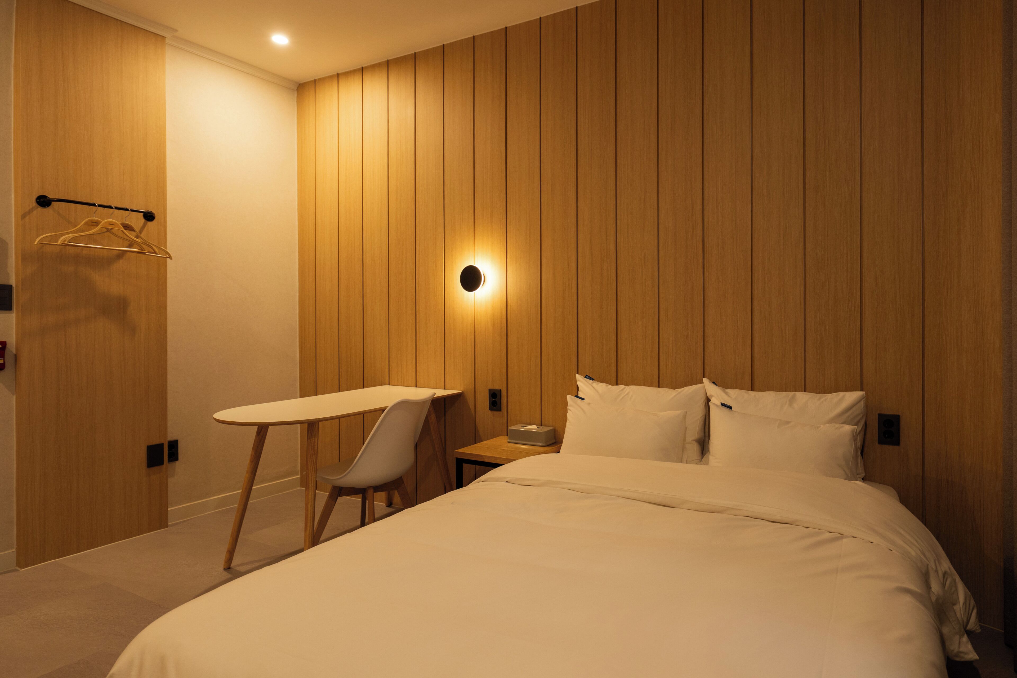 Quarto de hotel com cama, escrivaninha, cadeira e cabideiro.