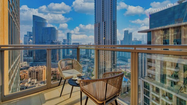 Varanda com cadeiras de vime e uma pequena mesa, com vista para um horizonte urbano com edifícios modernos e céu claro.