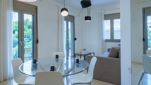 Uma sala de estar moderna com uma mesa de jantar de vidro, cadeiras brancas, um sofá e uma mesa de centro. Há duas luminárias pendentes acima da mesa e uma janela grande com cortinas.