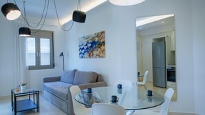 Uma sala de estar moderna com uma mesa de jantar de vidro, cadeiras brancas, um sofá cinza e um quadro na parede.