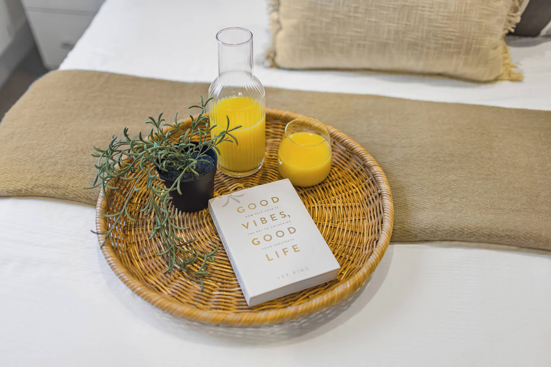 Bandeja de café da manhã com um livro intitulado "Good Vibes, Good Life" de Vick King, um copo de suco de laranja, outro copo de suco de laranja e uma planta em vaso sobre uma cama com travesseiro bege.