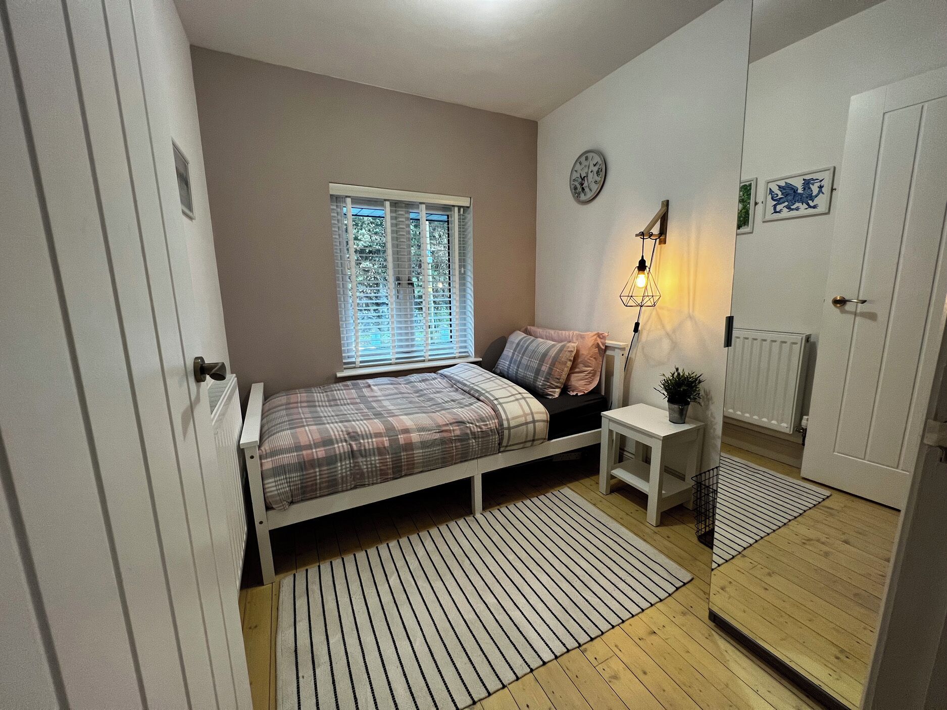 Quarto com cama, mesa de cabeceira, abajur, janela com persianas, radiador e porta.