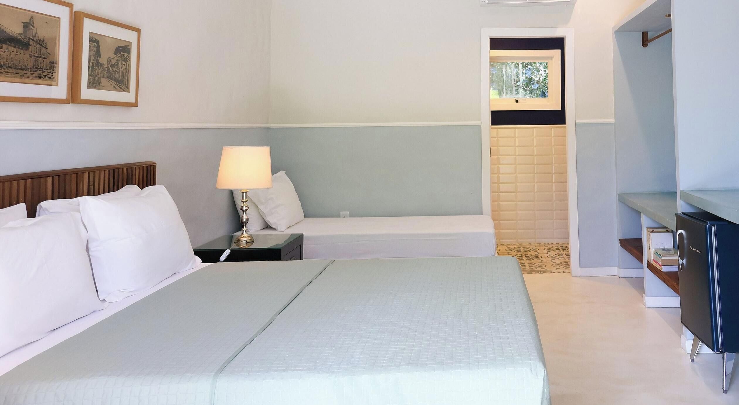 Quarto de hotel com uma cama grande, uma mesa de cabeceira com abajur, uma janela pequena e uma porta para um banheiro.