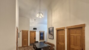Uma área de refeições com uma mesa grande, cadeiras e um lustre. Há portas de madeira e uma janela com vista.