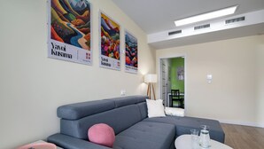 Uma sala de estar moderna com um sofá cinzento, uma pequena mesa branca e uma almofada rosa. Dois controles remotos estão sobre uma bandeja rosa.