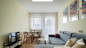 Uma sala de estar moderna com um sofá cinzento, uma TV sobre um móvel, uma área de refeições com cadeiras e uma mesa, e uma janela grande com cortinas.