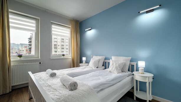 Uma cama de casal com roupa de cama branca, duas mesas de cabeceira com abajures, uma janela com cortinas e vista para prédios.