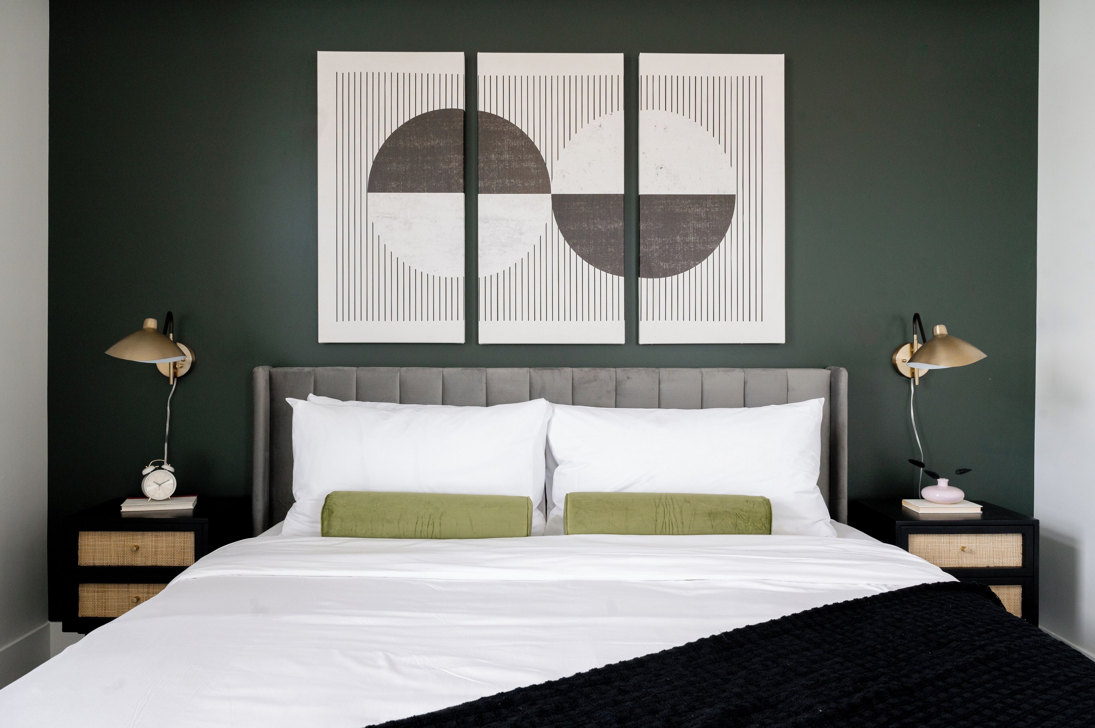 Cama bem arrumada com almofadas brancas e verdes, cabeceira estofada cinza e manta preta. Há duas mesas de cabeceira com abajures e uma obra de arte na parede sobre a cama.