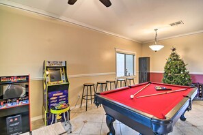 Uma mesa de bilhar com superfície em feltro vermelho, um taco e um suporte para bolas. Ao fundo, um jogo de arcade Midway Mortal Kombat e uma árvore de Natal.