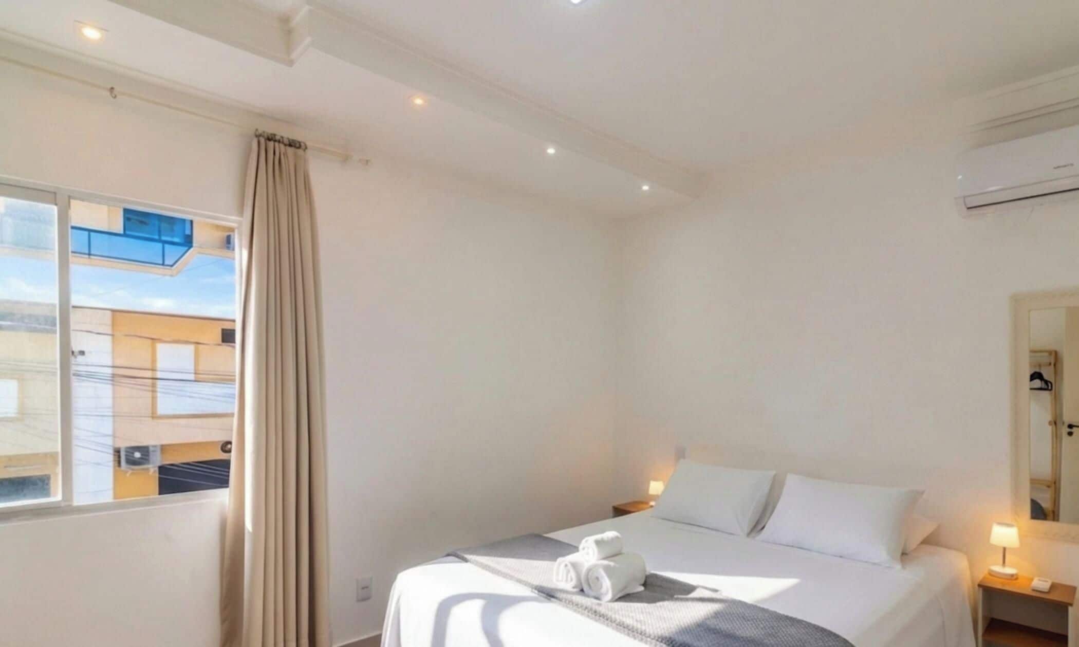 Quarto de hotel com cama, abajures, janela com cortinas e aparelho de ar condicionado.