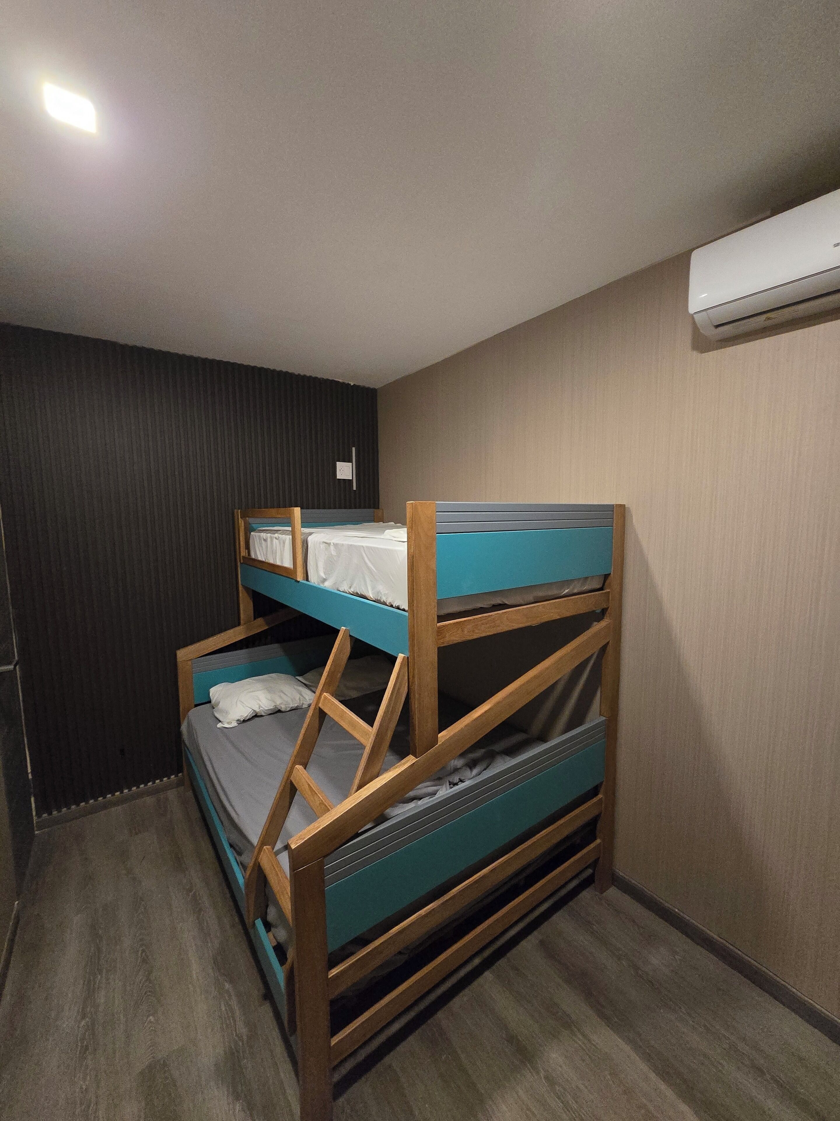 Quarto com beliche de madeira, colchão branco na parte superior e colchão cinza na inferior. Ar condicionado fixado na parede à direita e uma tomada na parede.