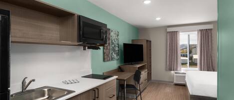 Quarto de hotel compacto com cozinha compacta, televisão de tela plana, cama e janela com cortinas.