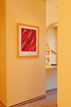Um corredor com uma pintura abstrata emoldurada na parede, piso de madeira e um espelho redondo que reflete parte do quarto.
