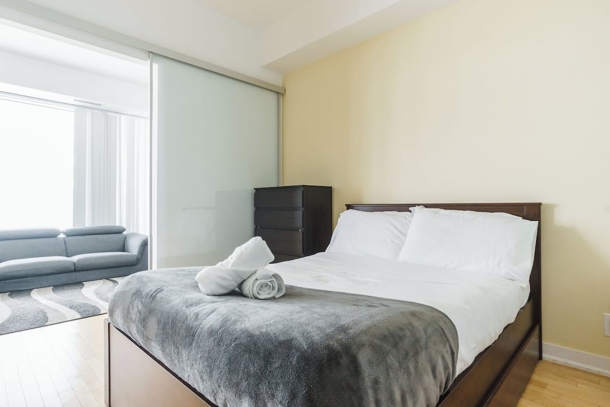 Quarto de hotel com cama, sofá cinza, armário de madeira e uma janela grande.