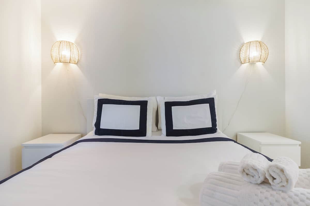 Uma cama bem arrumada com roupa de cama branca e azul-marinho, duas luminárias de cabeceira e uma mesa de cabeceira branca.
