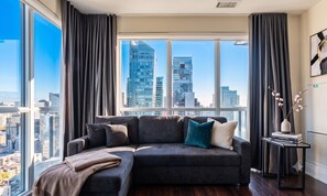 Uma sala de estar moderna com um sofá seccional, uma mesa lateral e vista para o horizonte urbano através de amplas janelas.