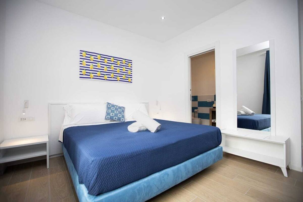Quarto de hotel moderno com cama grande, espelho e banheiro visível através de uma porta aberta.