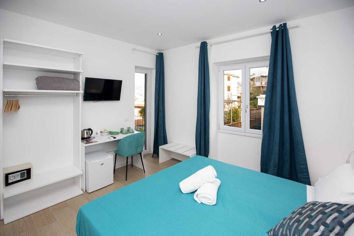 Quarto de hotel com cama, escrivaninha, televisão e janela com cortinas.