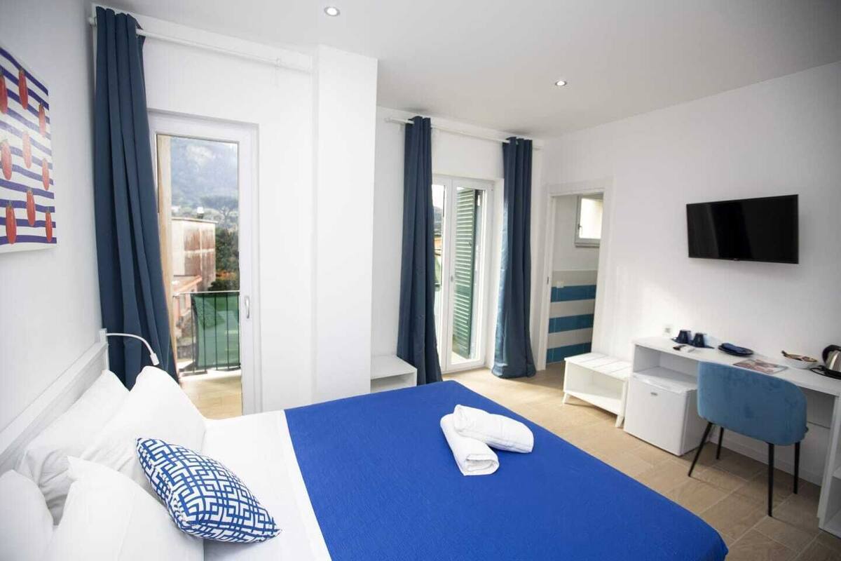 Quarto de hotel com cama, escrivaninha, televisão e varanda com cortinas.