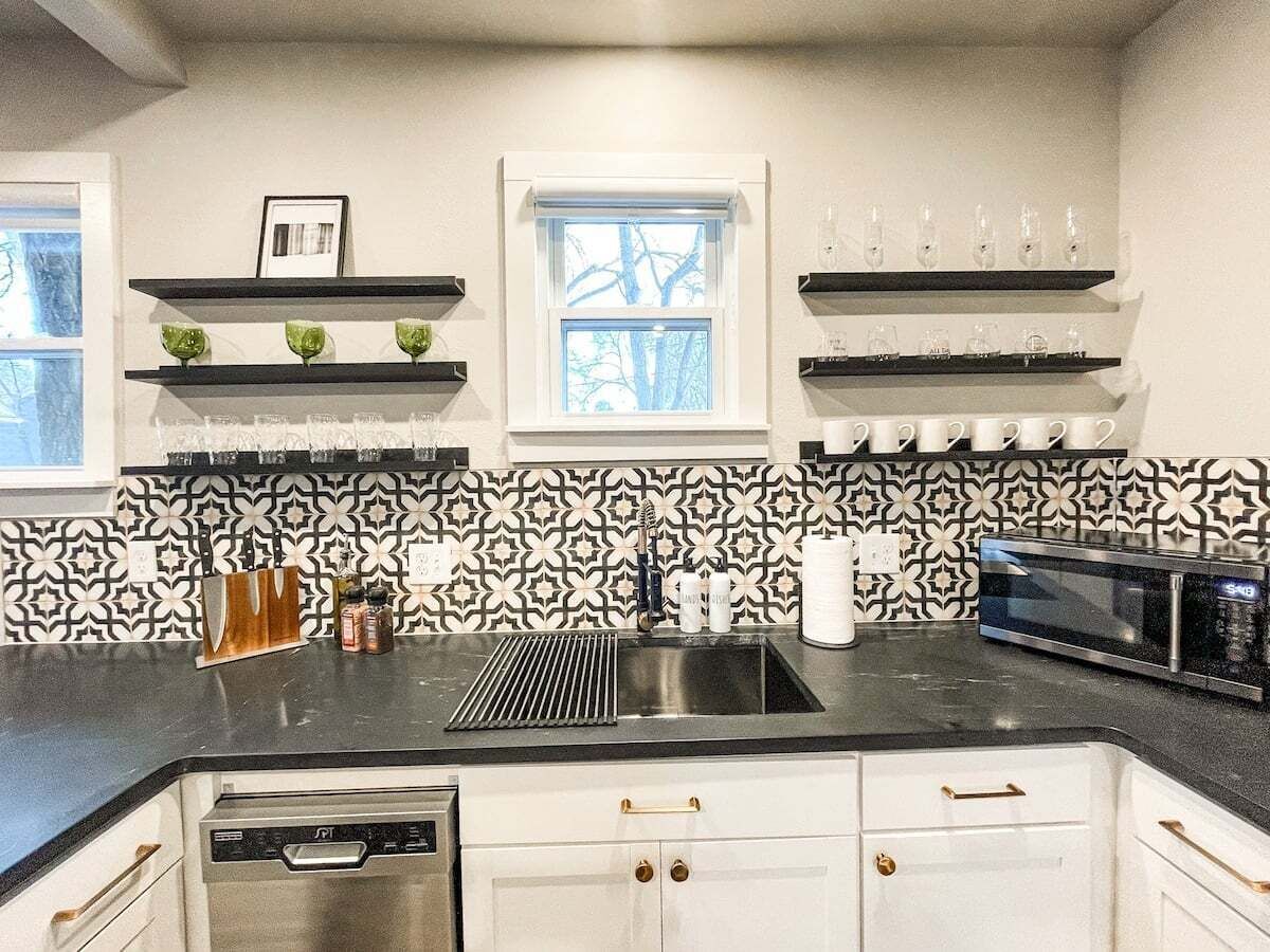 Uma cozinha com bancada preta, armários brancos, lava-louças de aço inoxidável, micro-ondas e janela com persianas.