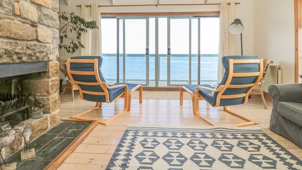 Uma sala de estar acolhedora com parede de pedra, duas poltronas de madeira, lareira e vista para o mar através de grandes portas de vidro.